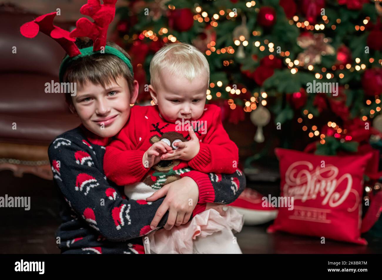 A smiling brother in a Christmas outfit holds his little sister near a ...
