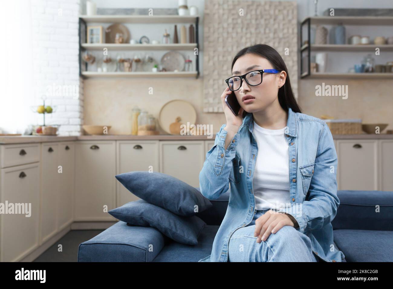 Worried and confused young Asian woman sitting on sofa at home and ...