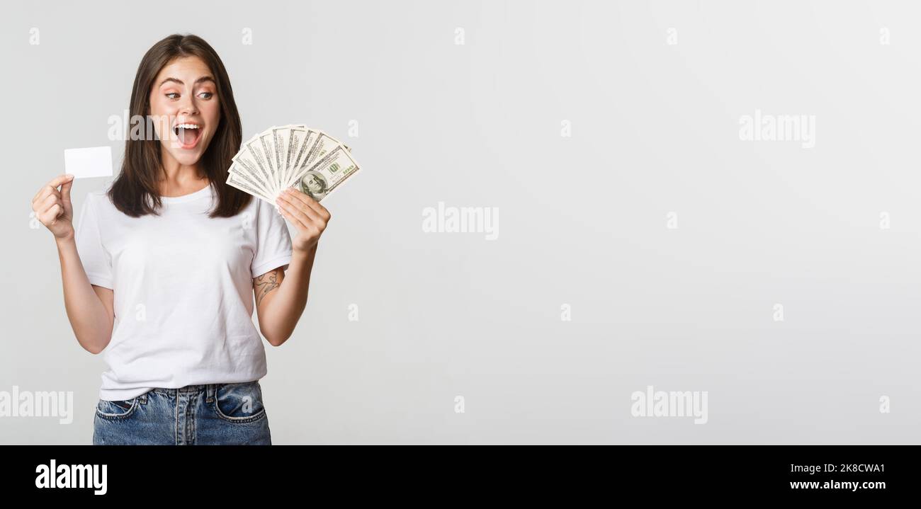 Amused beautiful girl looking at money and holding credit card ...