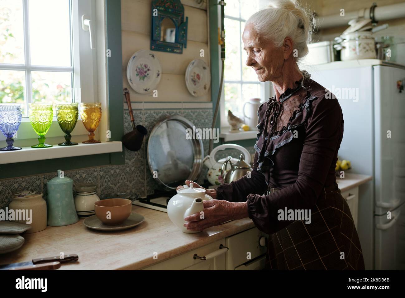 Serene senior woman holding teapot while brewing tea for her guests or ...