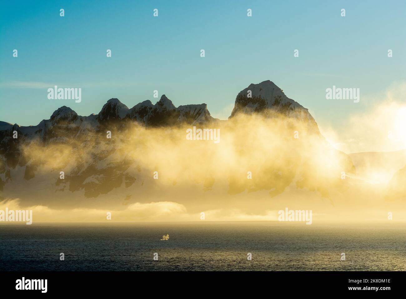 early morning sun lights up cloudy fief mountains peaks on wiencke island with whale blow in ...
