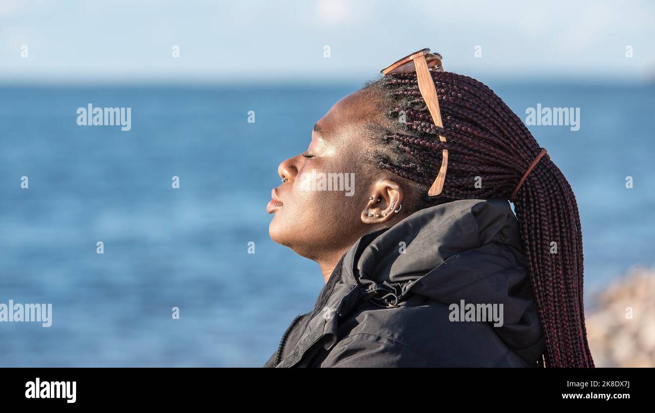 African woman sits by the Swedish Baltic Sea and enjoys the sunset ...