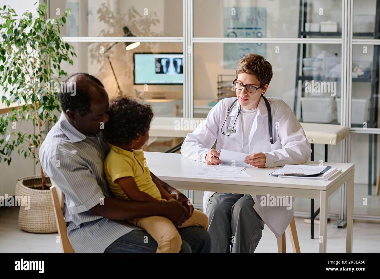 Female doctor sitting at table and talking to child while he sitting on ...
