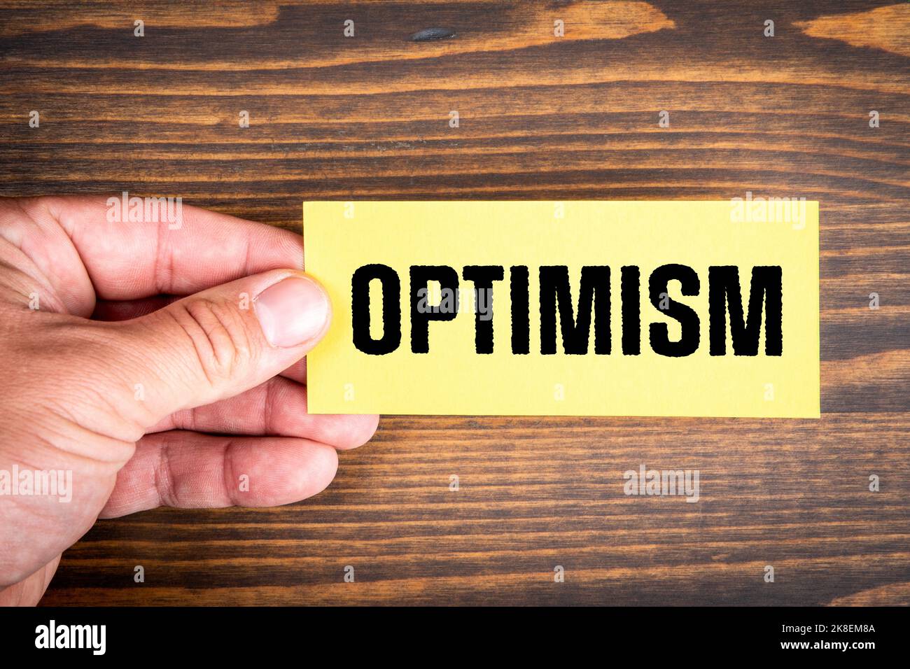 OPTIMISM. Piece of note paper with text in a man's hand. Wood texture ...