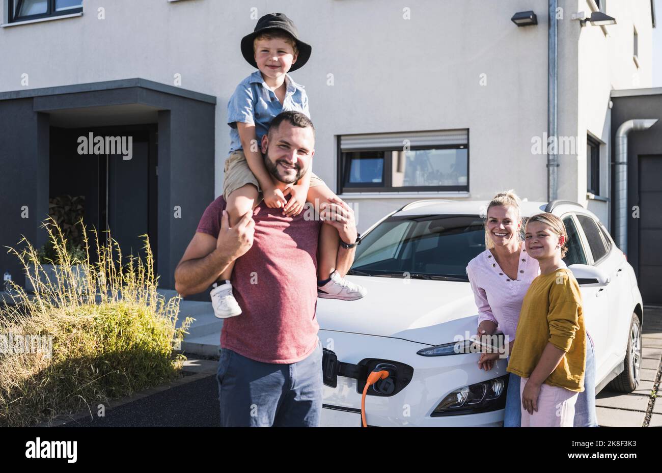 Happy family standing in front of family home and charging electric car ...