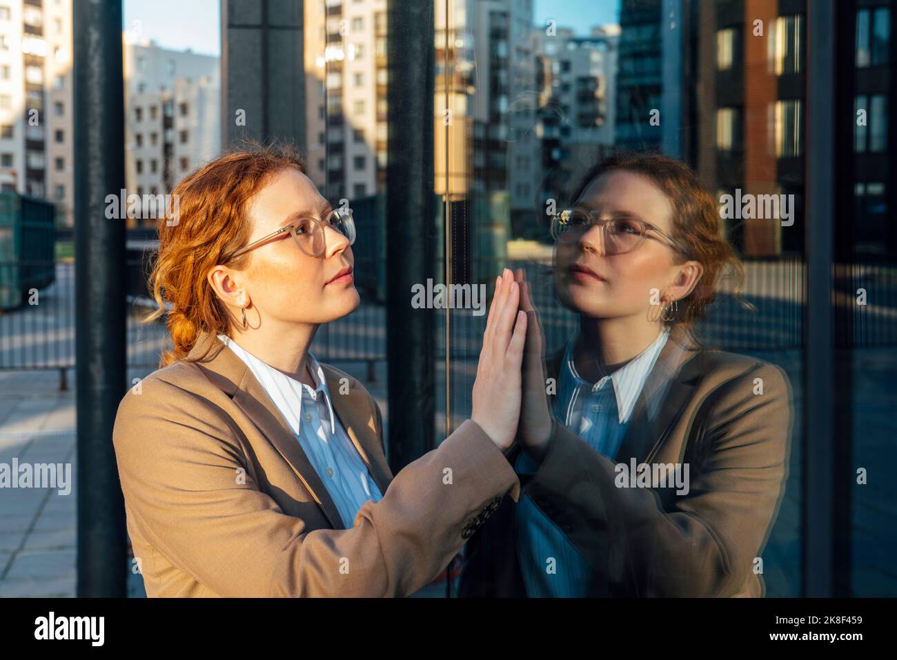 Thoughtful young redhead businesswoman touching reflection on glass ...