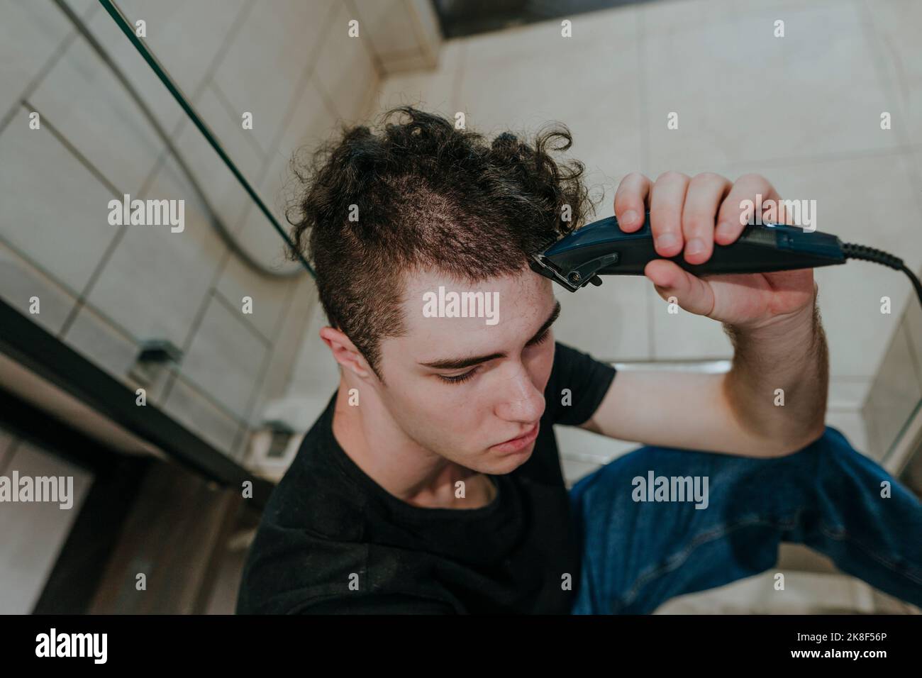 Sad young man with electric razor shaving his own hair in bathroom ...