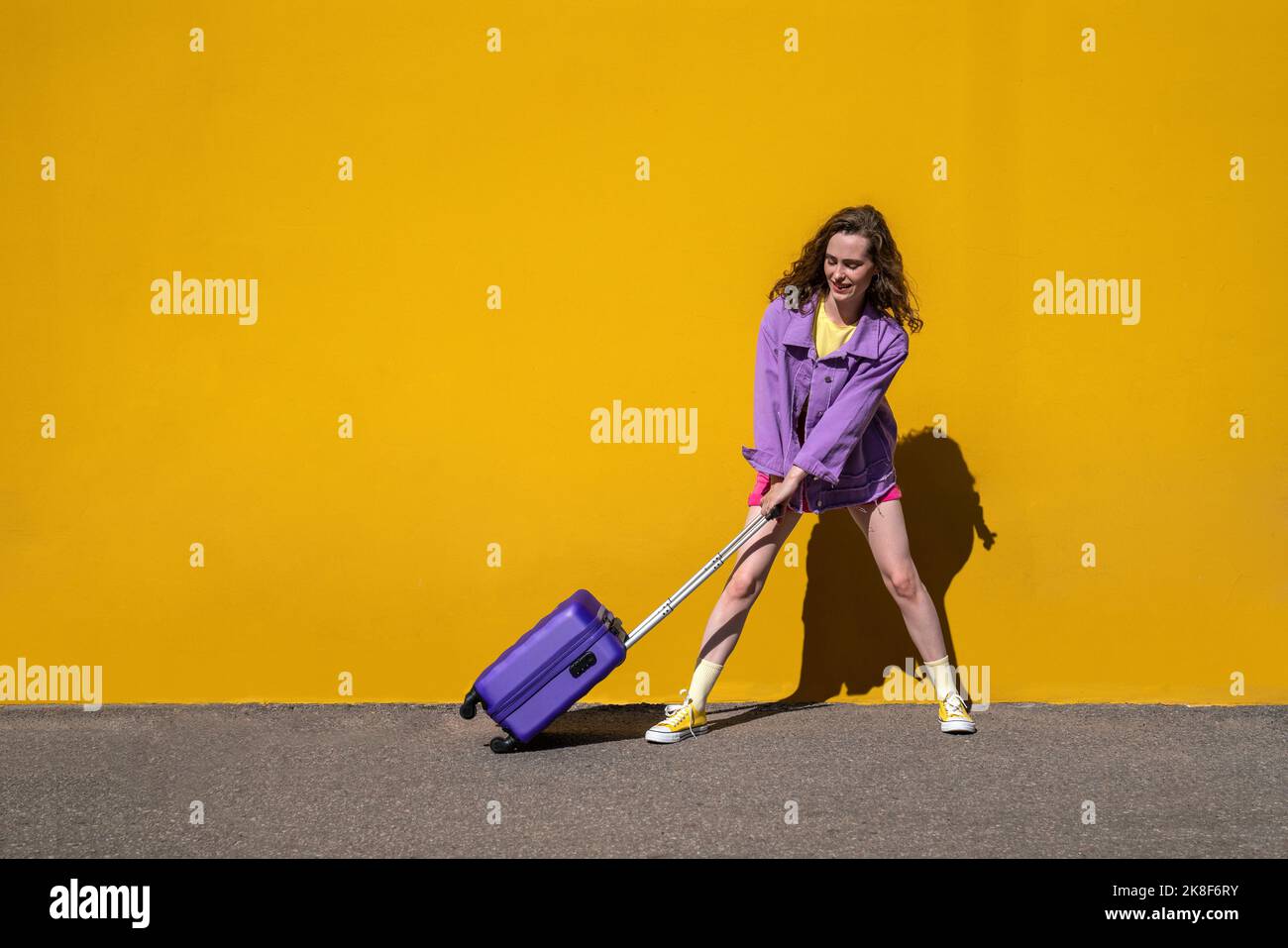 Woman pulling wheeled luggage in front of yellow wall Stock Photo - Alamy