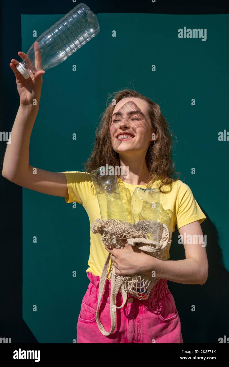 Smiling woman shielding eyes holding mesh bag and plastic bottle on ...