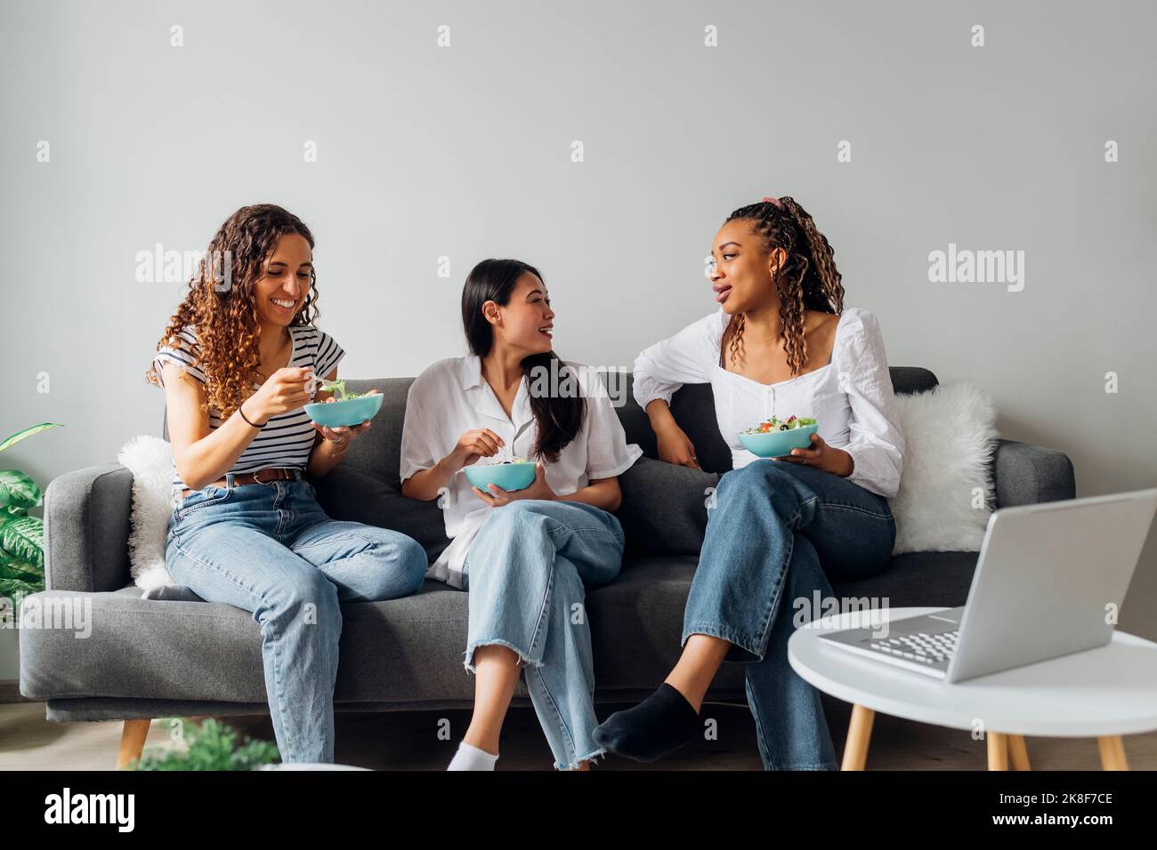 Smiling young multiracial roommates eating salad together sitting on ...