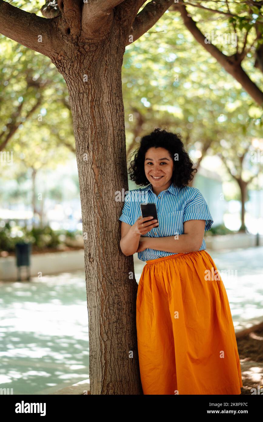 Smiling woman holding mobile phone leaning on tree trunk Stock Photo ...