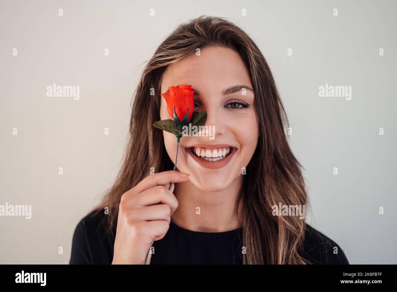 Happy young woman covering eye with red rose against white background ...
