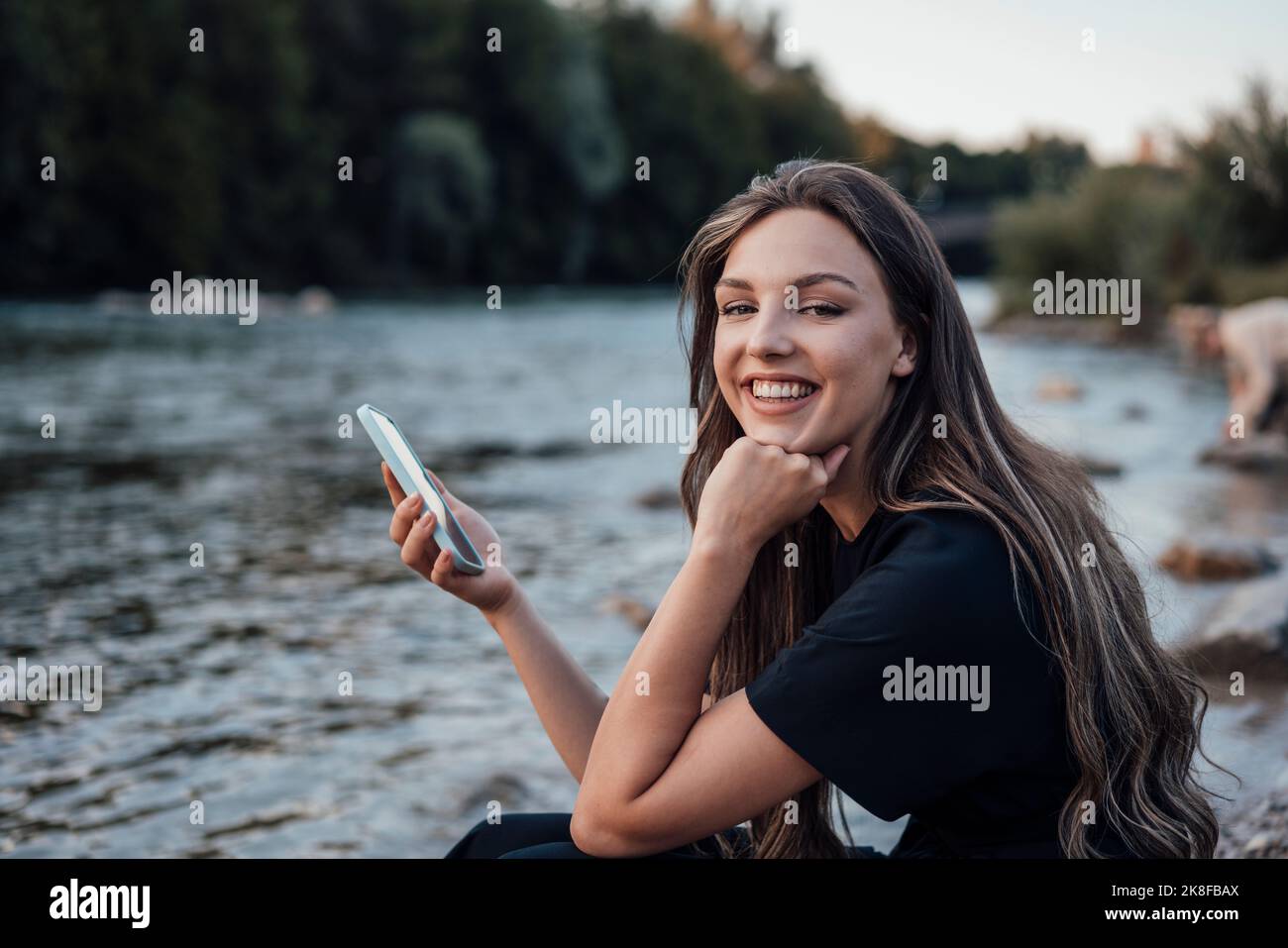 Happy young woman holding mobile phone sitting with hand on chin at ...
