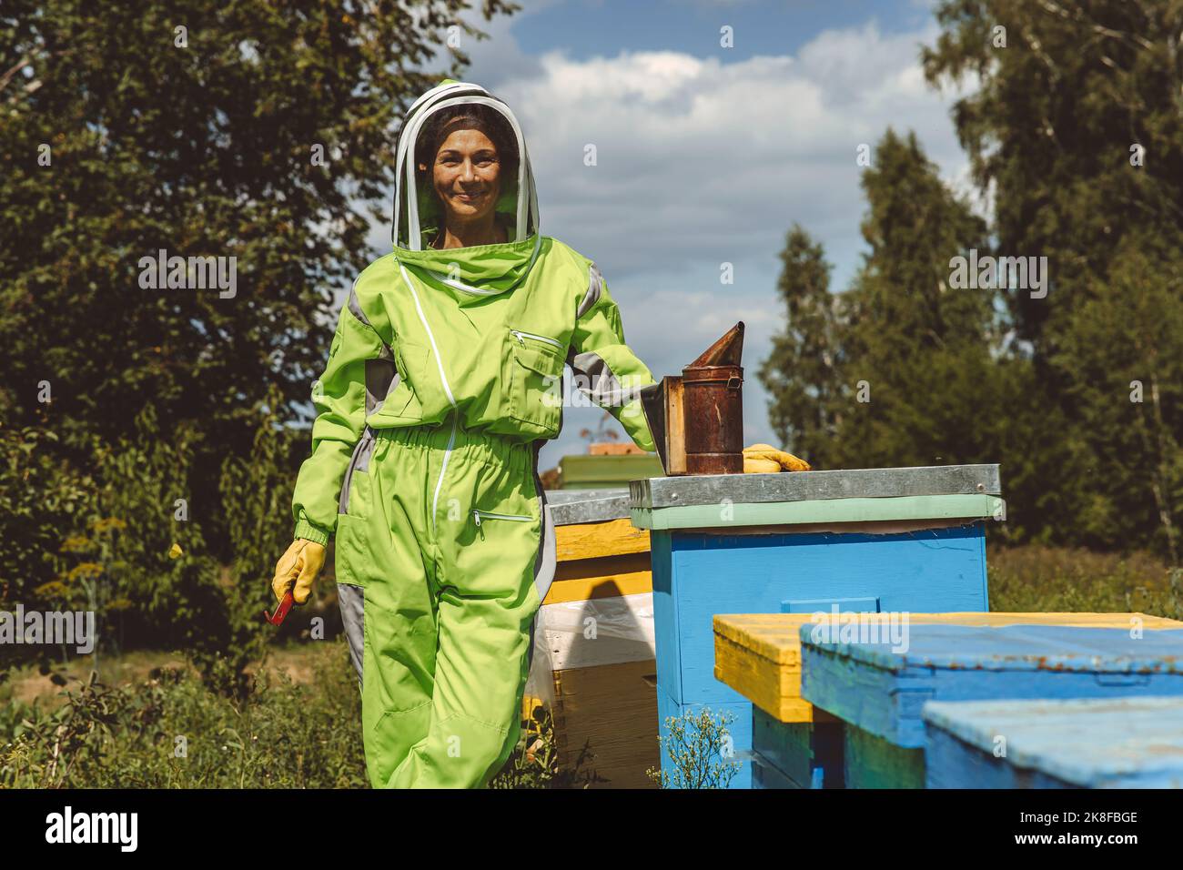 Smiling beekeeper with bee smoker standing by wooden boxes on sunny day ...