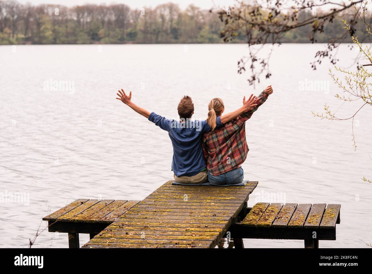 Carefree couple with arms outstretched sitting on jetty at lake Stock ...