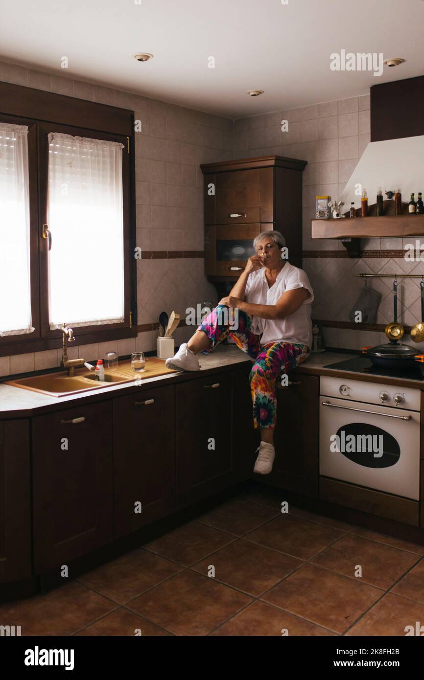Woman smoking cigarette sitting on kitchen counter at home Stock Photo ...