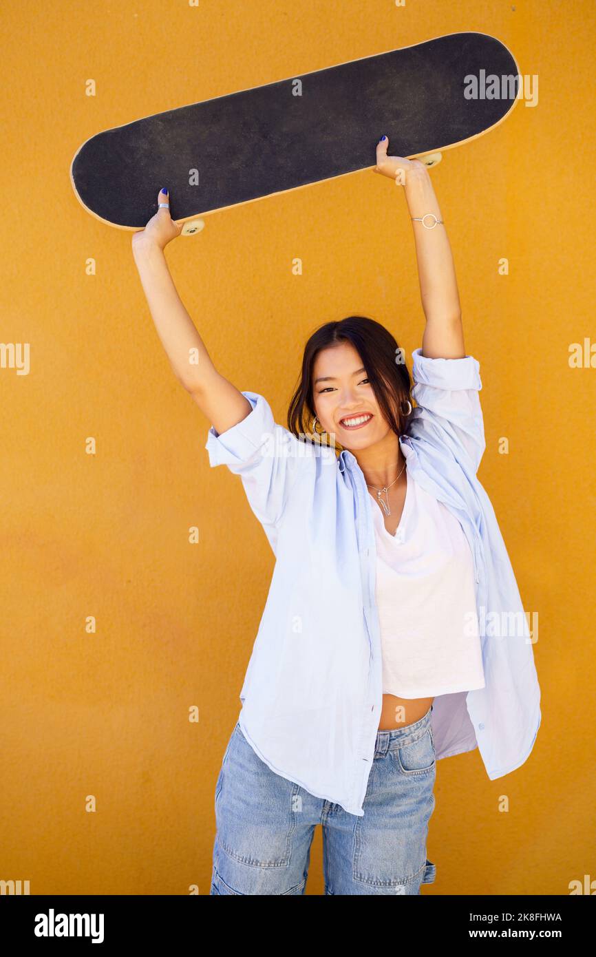 Happy young woman holding aloft skateboard in front of yellow wall ...