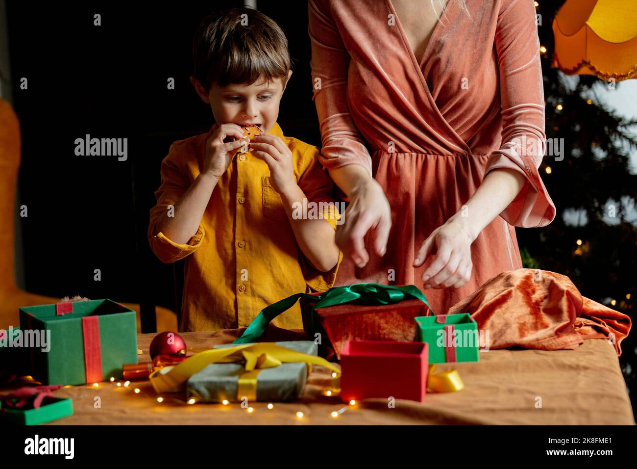 Boy eating snack with mother wrapping gifts on table at home Stock ...