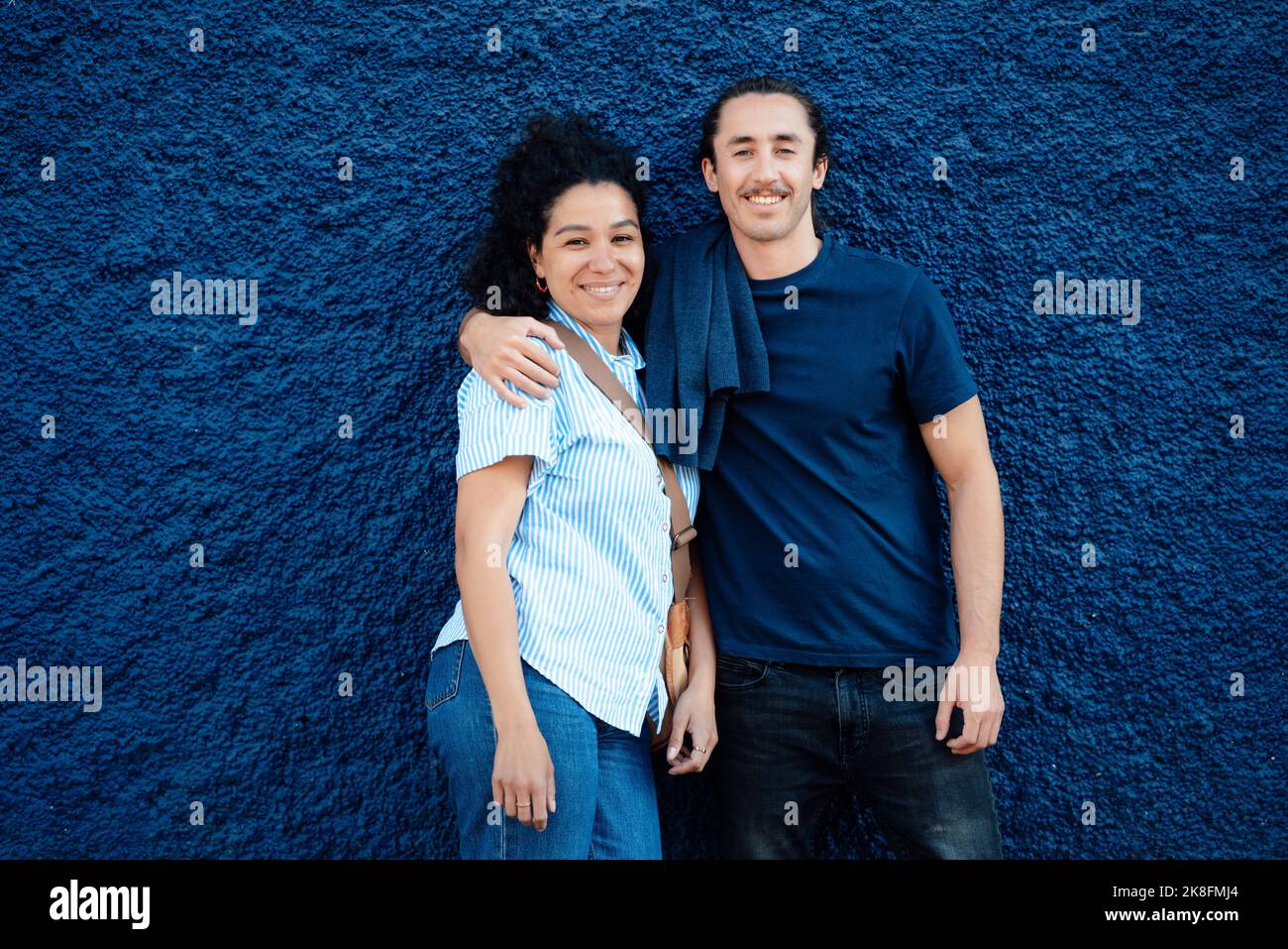 Smiling boyfriend with arm around of girlfriend in front of blue wall ...