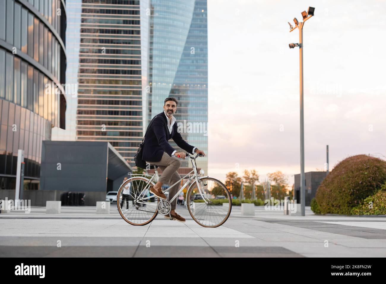 Mature commuter riding bicycle in front of office building at sunset ...