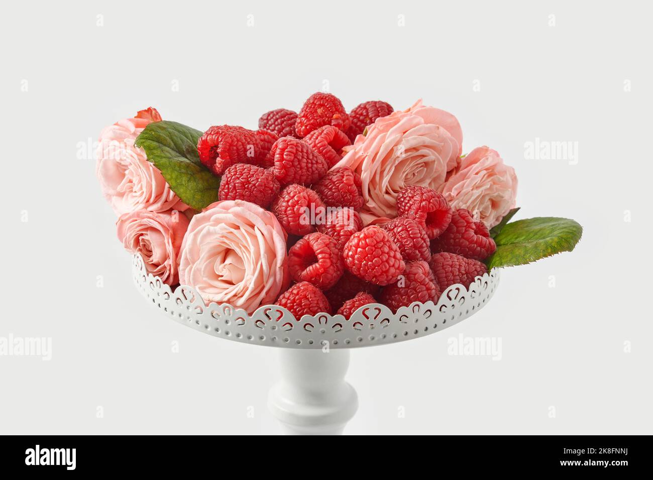 Fresh raspberries with pink roses on pedestal cakestand against white ...