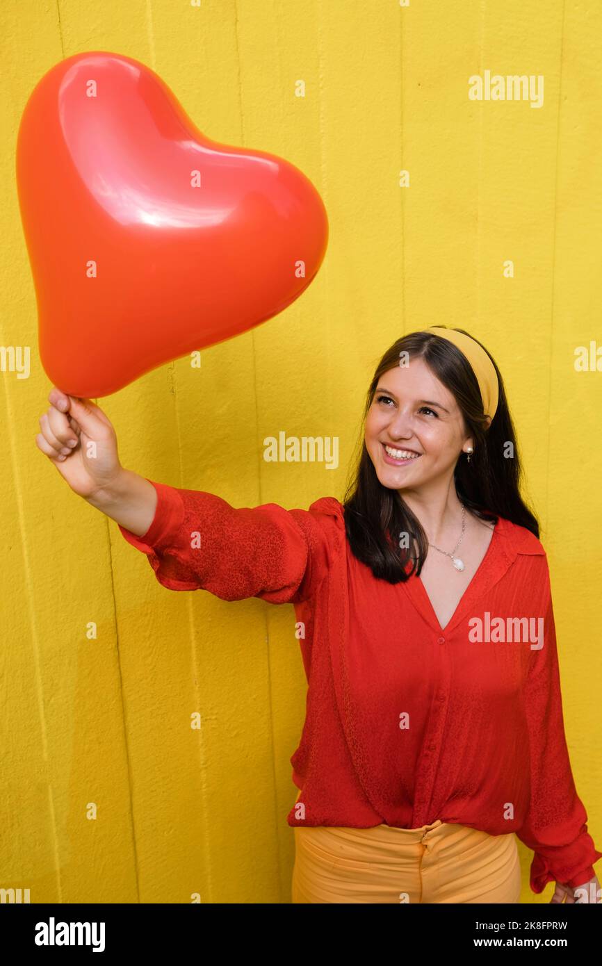 Happy woman holding heart shape balloon in front of yellow wall Stock ...