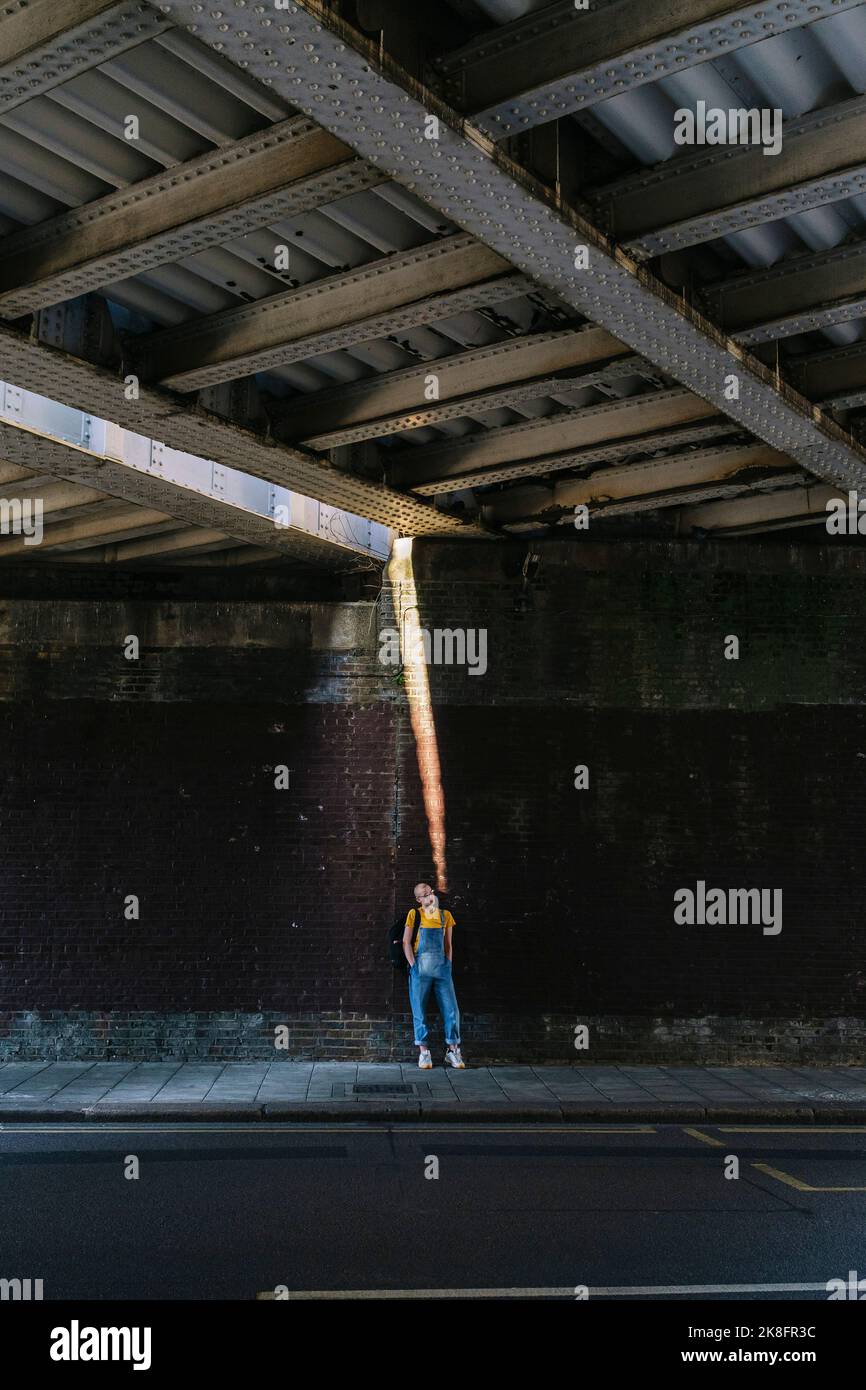 Non-binary person standing in front of wall below bridge Stock Photo ...