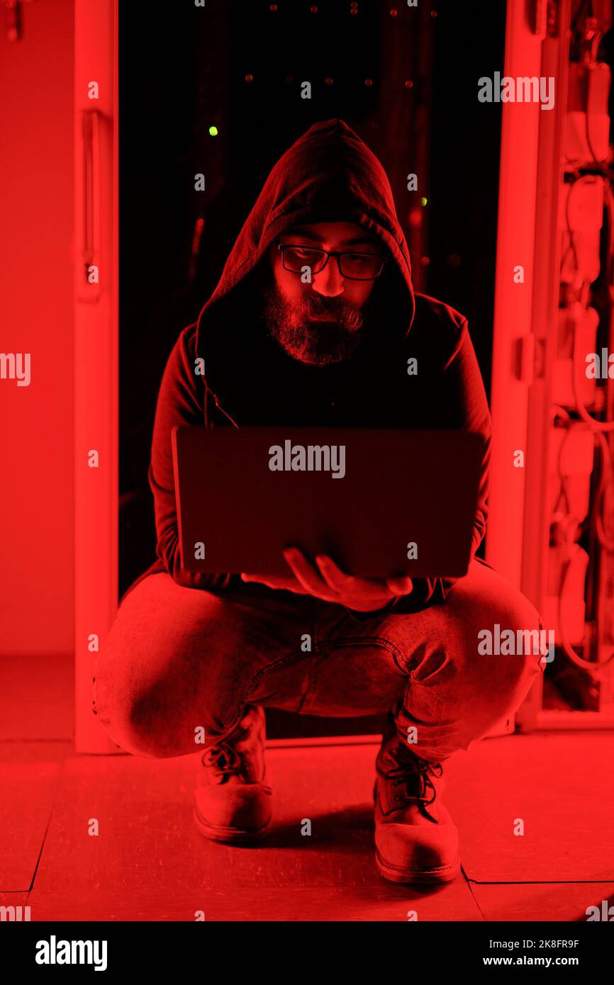 Computer hacker with laptop crouching in server room with red light Stock Photo - Alamy