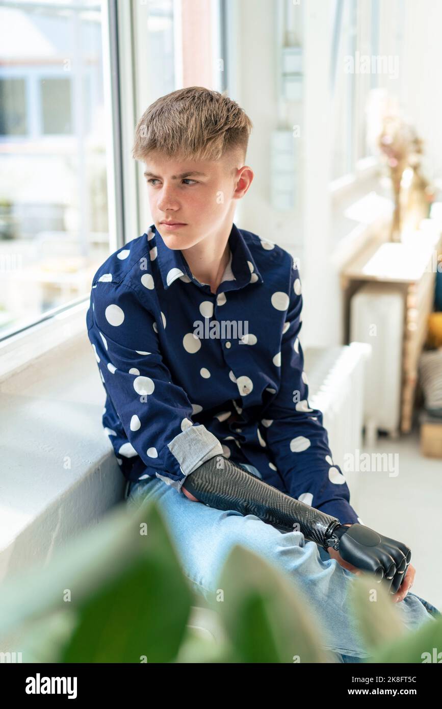 Thoughtful teenage boy with amputated arm looking out through window ...