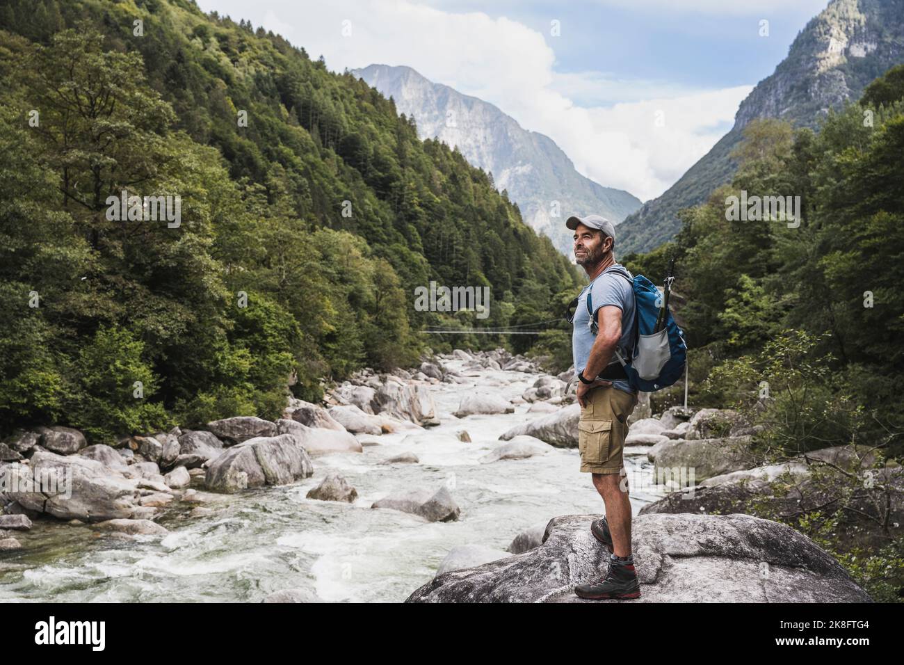 Mature man with hand on hip standing by river in front of mountains ...
