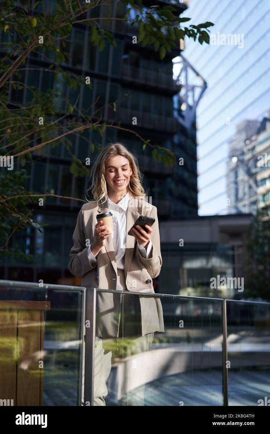 Corporate woman in suit, drinks coffee, holds mobile phone in hand ...