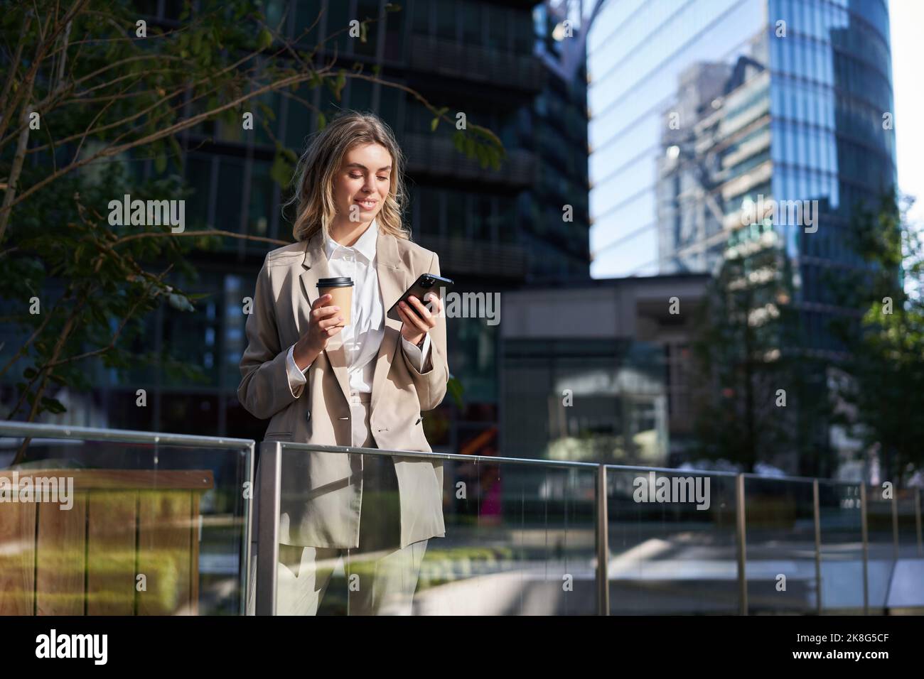 Corporate woman in suit, drinks coffee, holds mobile phone in hand ...