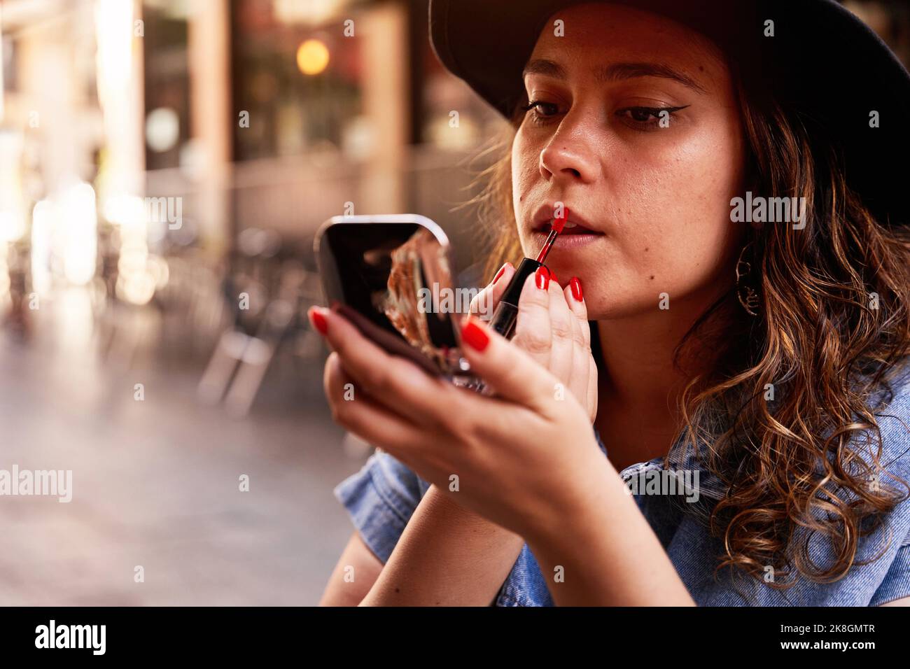 Young female in hat looking in mirror and smearing red lipstick on lips ...