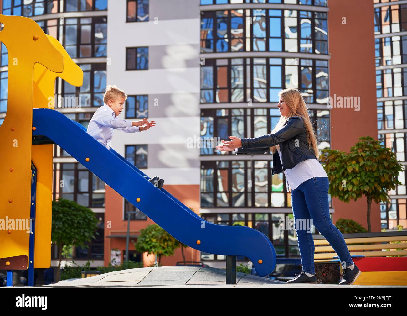 Pretty mum catching male kid, riding on slide at modern courtyard of ...