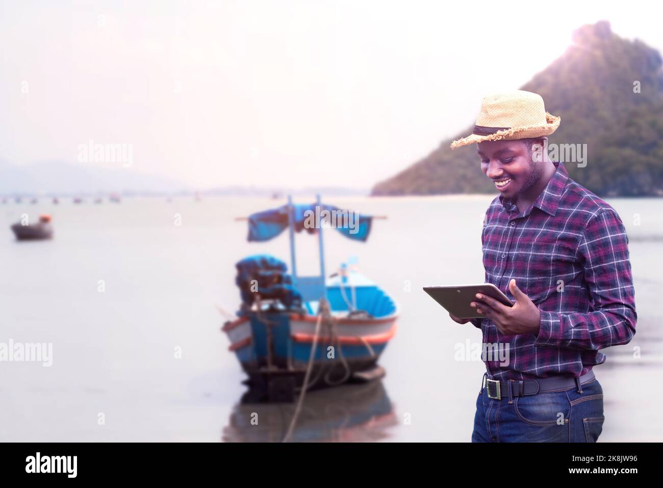 African trawler captain stands in front of the ship using a tablet to ...