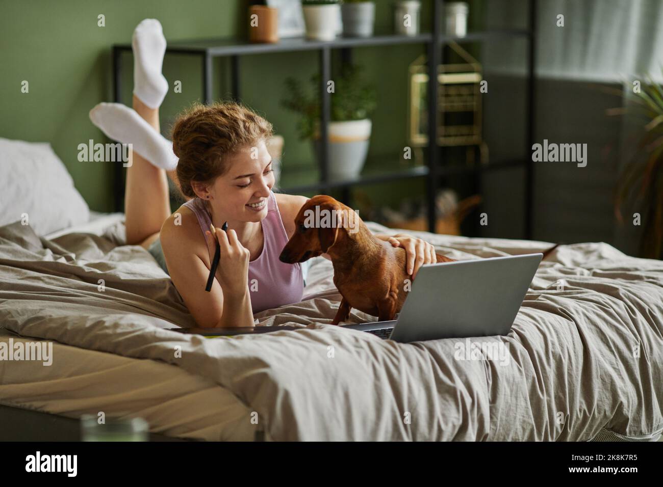 Young girl using laptop and playing with her dog while relaxing in bed ...