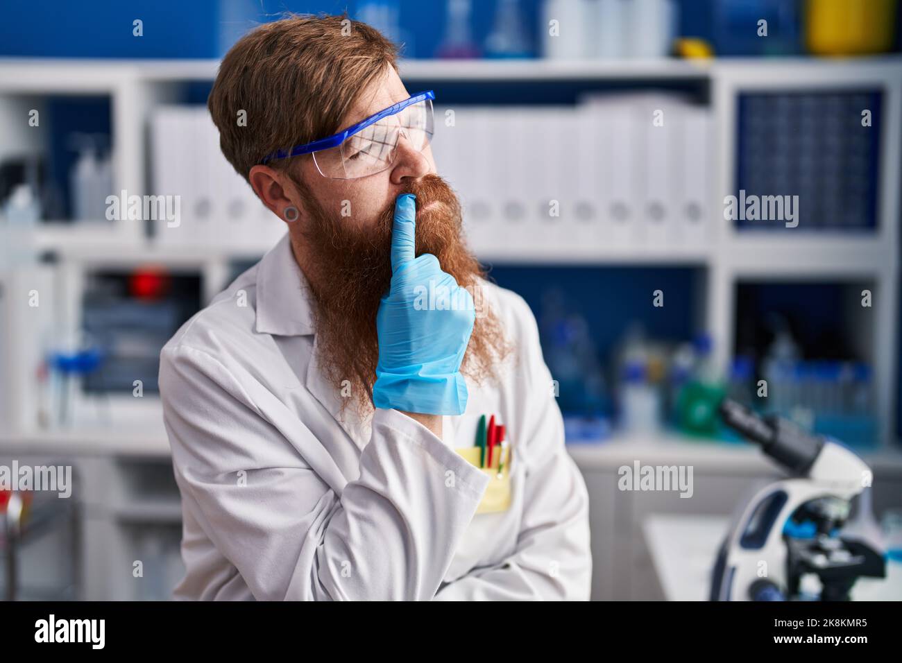 Young redhead man scientist sitting with doubt expression at laboratory ...