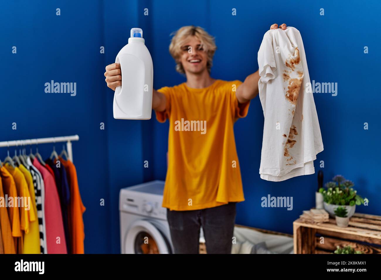 Young blond man holding dirty t shirt and detergent at laundry room ...