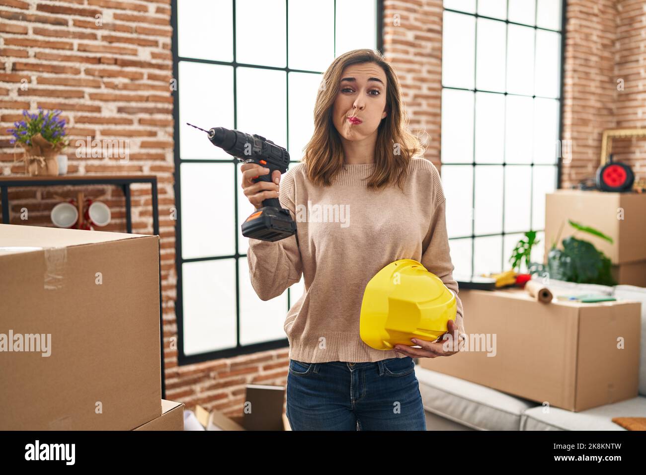 Young woman holding drill at new home puffing cheeks with funny face ...