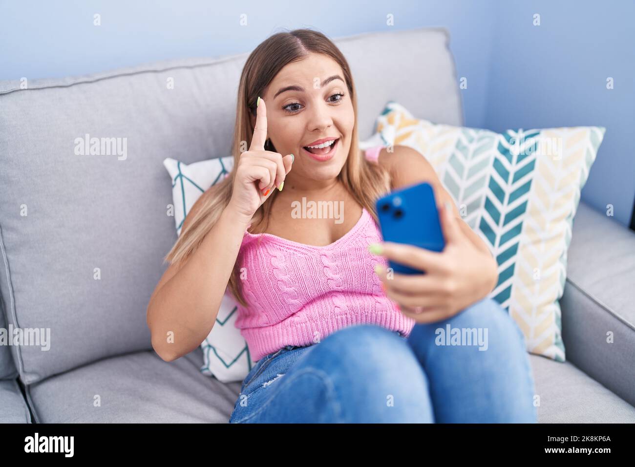 Young blonde woman doing video call with smartphone sitting on the sofa ...