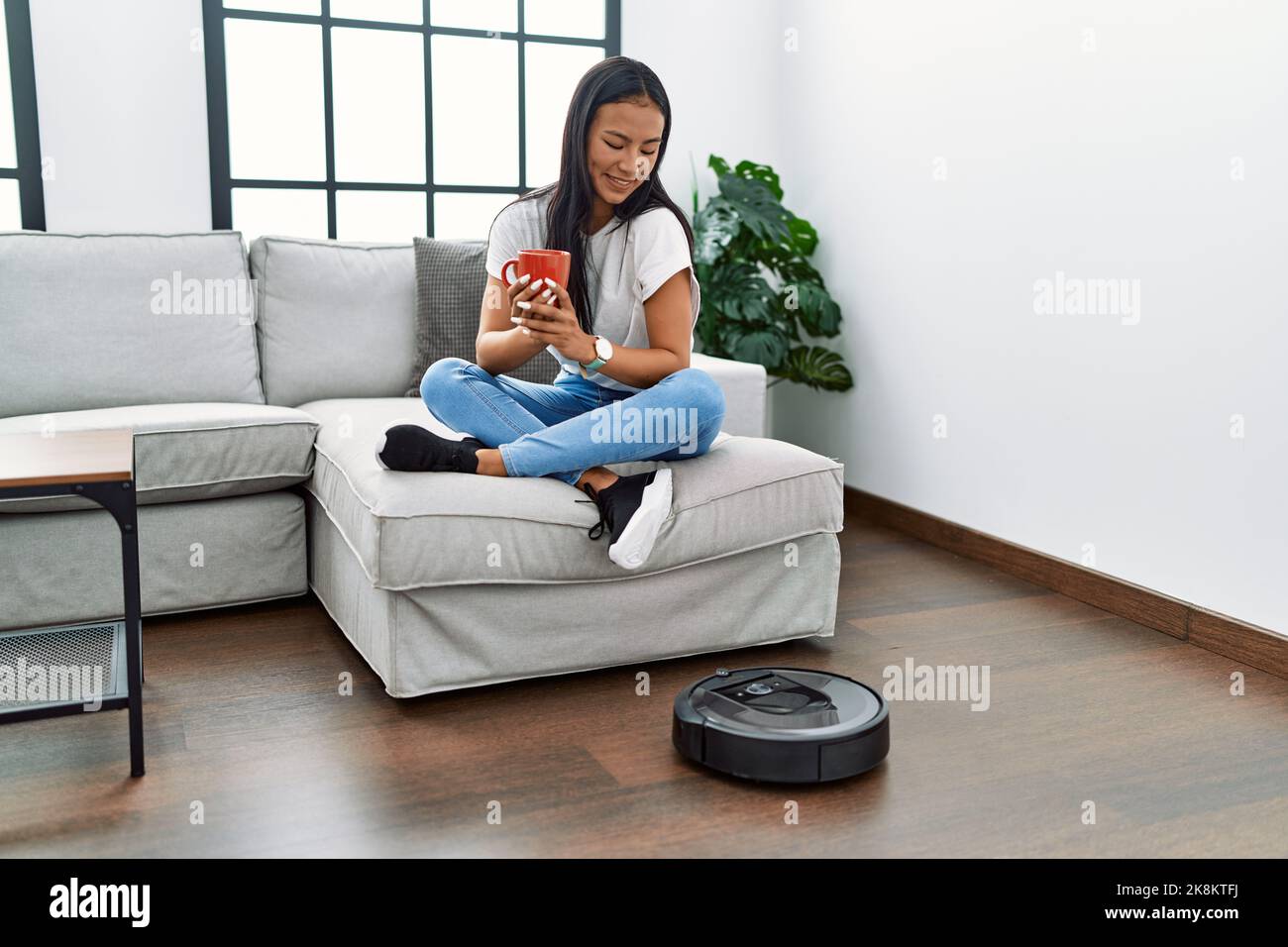 Young latin woman drinking coffee cleaning floor using smart robot ...