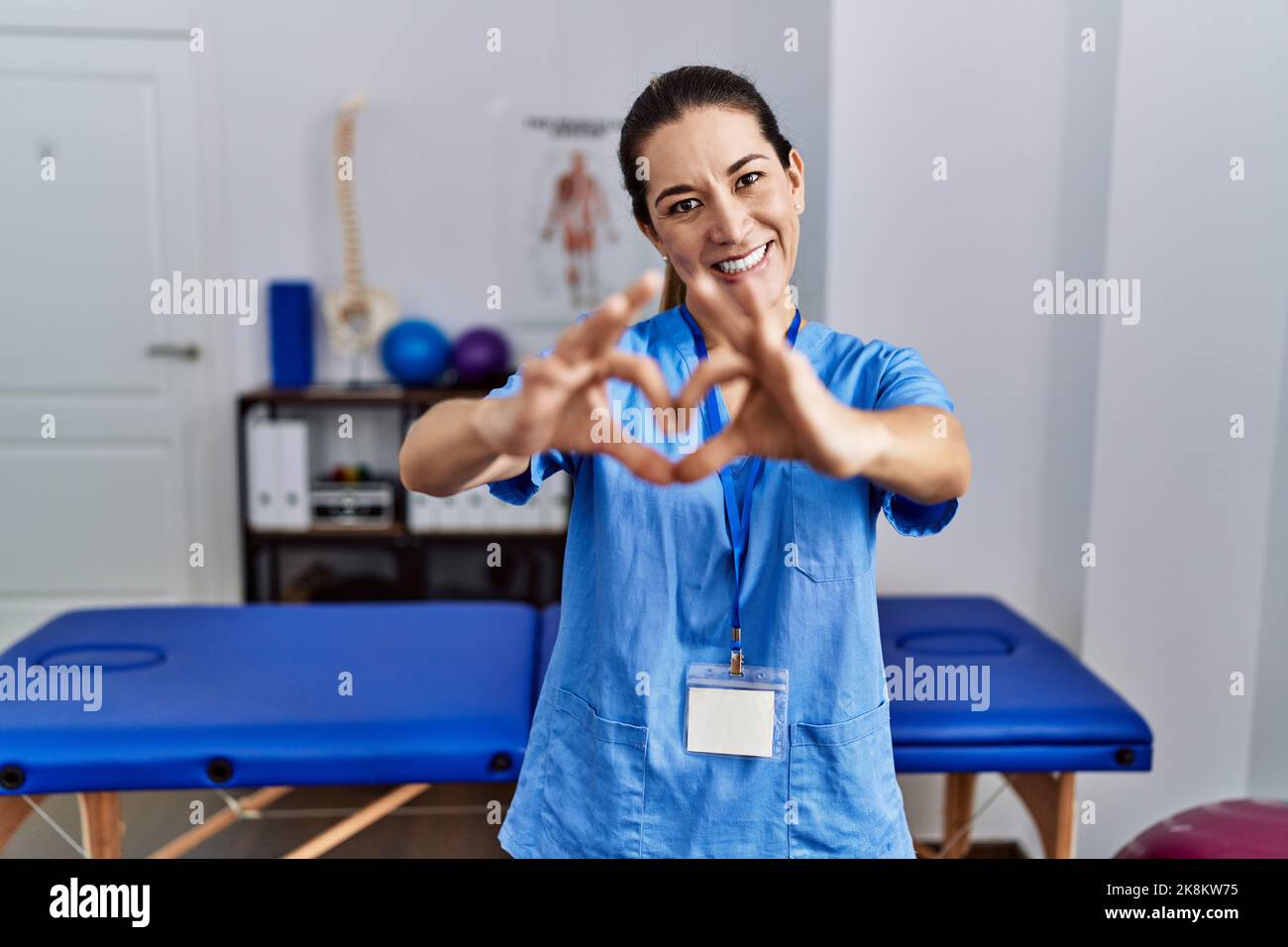 Young hispanic woman wearing physiotherapist uniform standing at clinic smiling in love doing ...