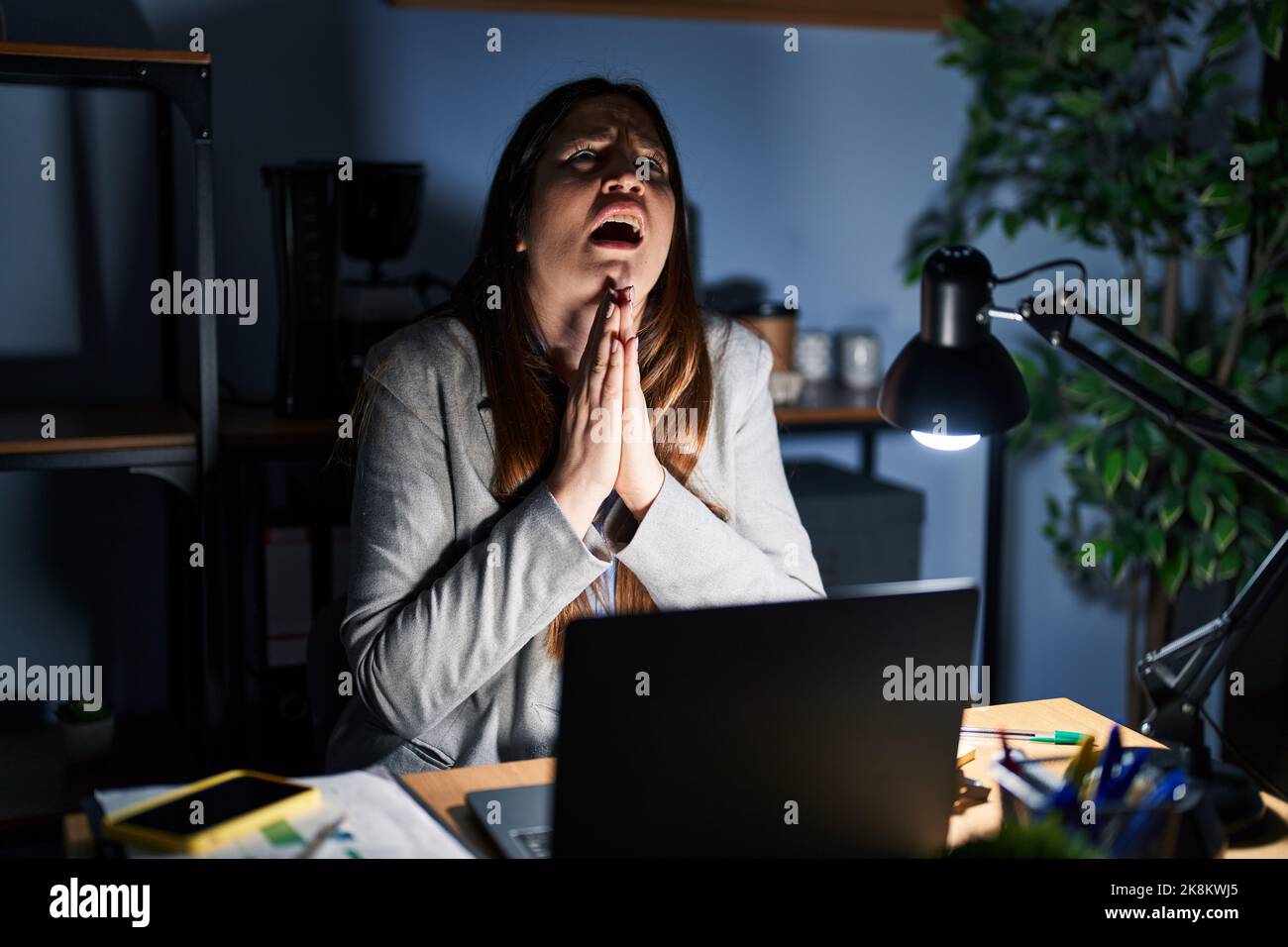 Young brunette woman working at the office at night begging and praying ...