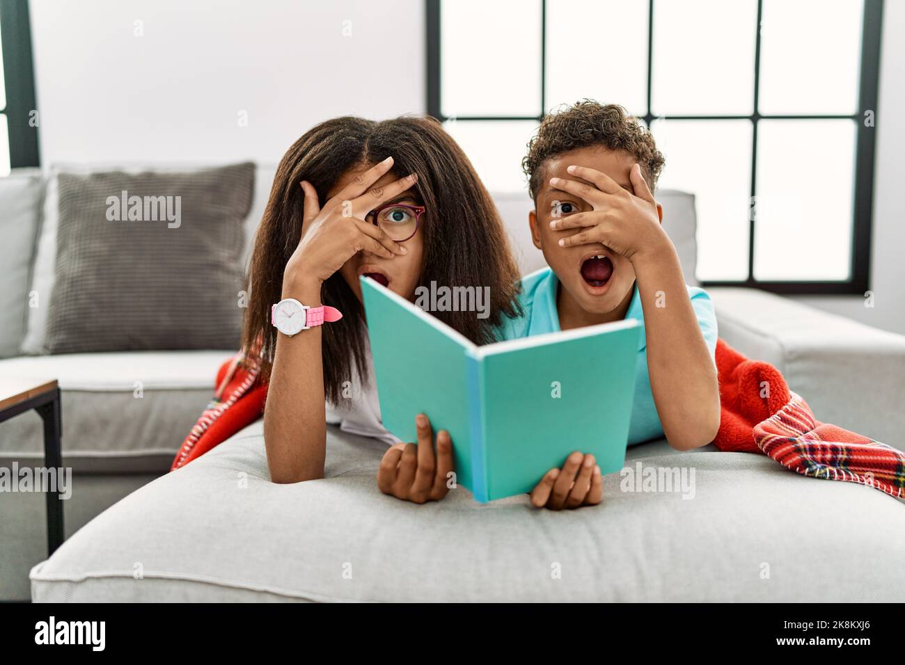 Two siblings lying on the sofa reading a book peeking in shock covering ...