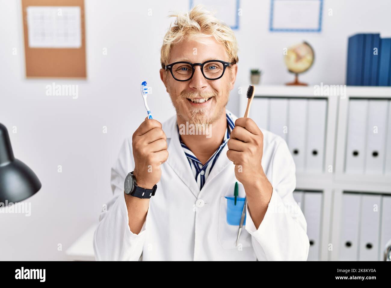 Young blond dentist man working at dentist clinic holding toothbrushes ...