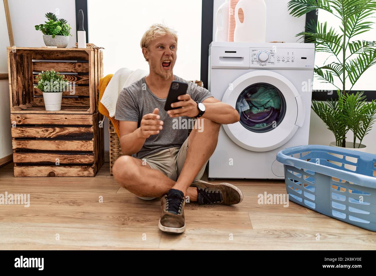 Young blond man doing laundry using smartphone angry and mad screaming ...