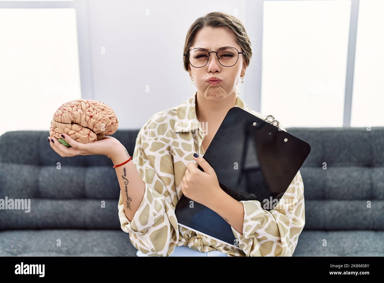Young hispanic woman holding brain working at psychology clinic puffing ...