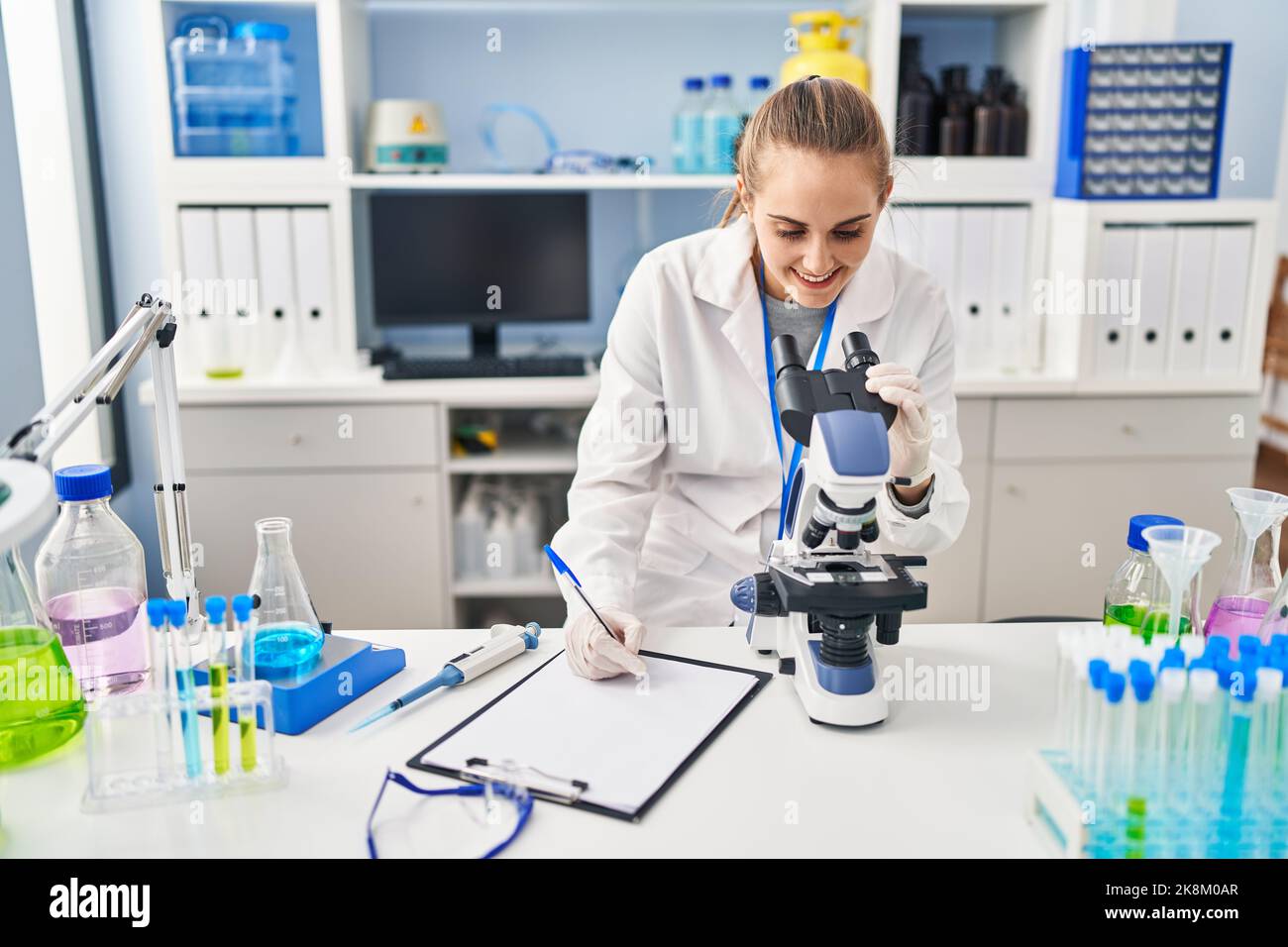 Young blonde woman wearing scientist uniform using microscope working ...