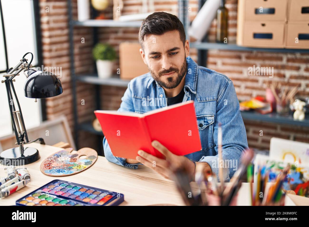 Young hispanic man reading book sitting on table at art studio Stock ...