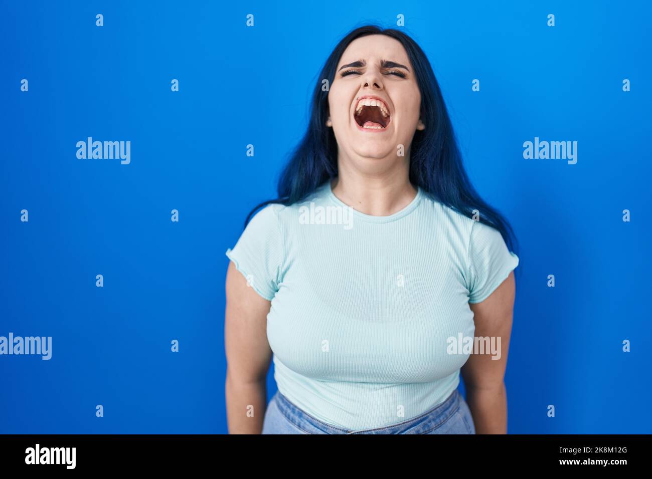 Young modern girl with blue hair standing over blue background angry ...
